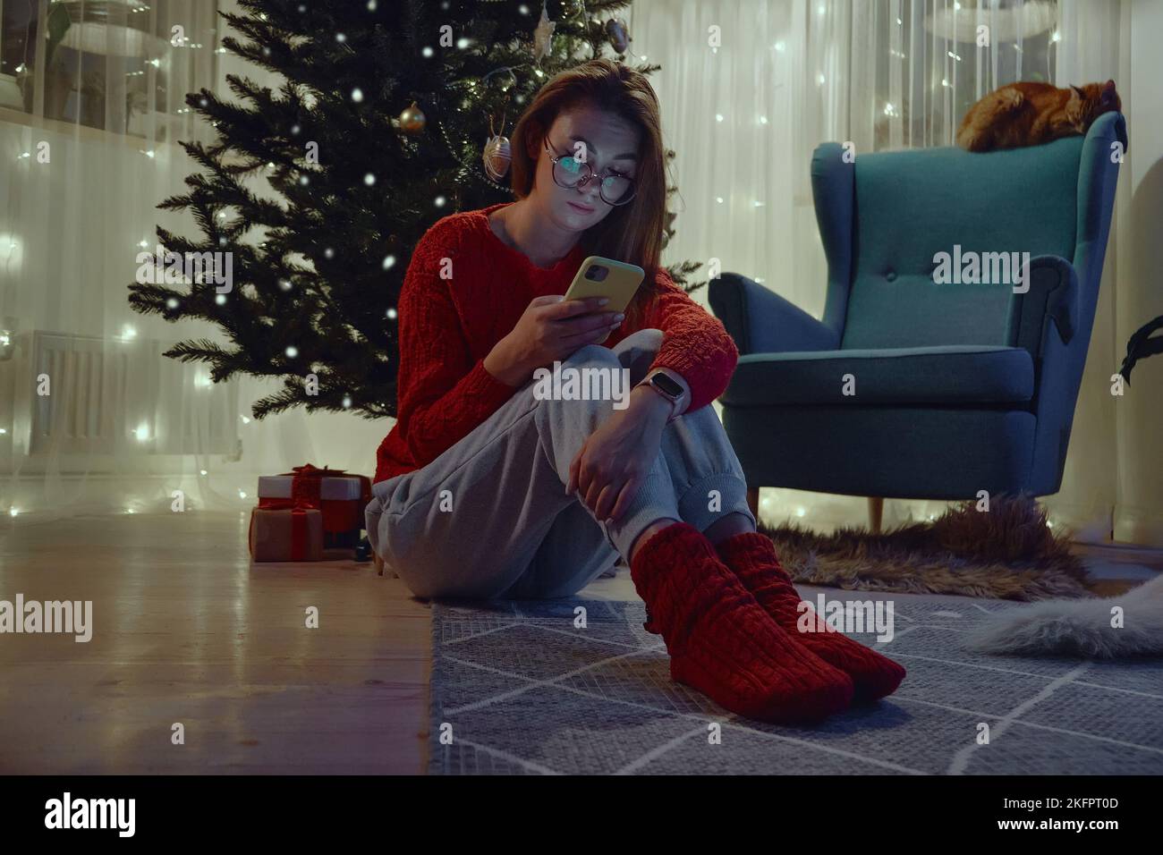 Woman using mobile phone sitting next to pet cat and christmas tree on ...