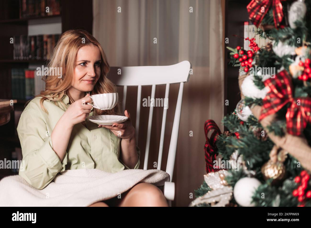 Young blonde woman sitting on a rocking chair next to a Christmas tree ...