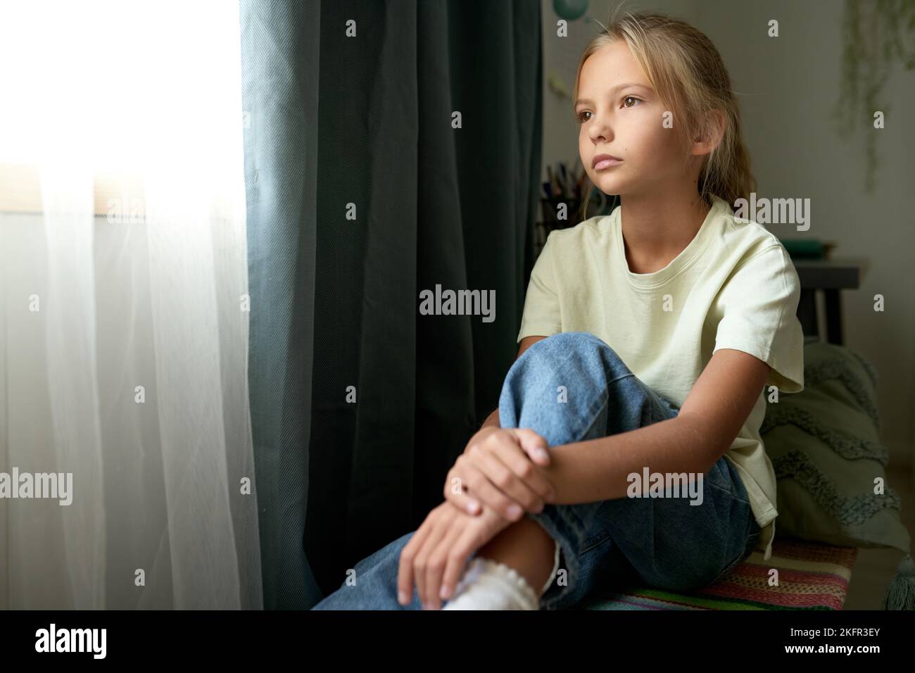 Sad little girl sitting alone in her room and looking at window with ...