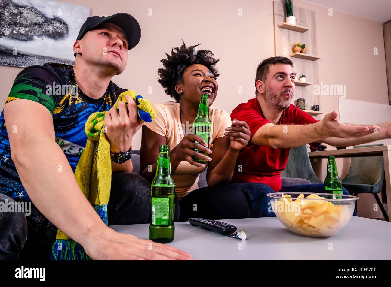 Friends watching a football match on TV, drinking beer and eating chips, cheering for their ...