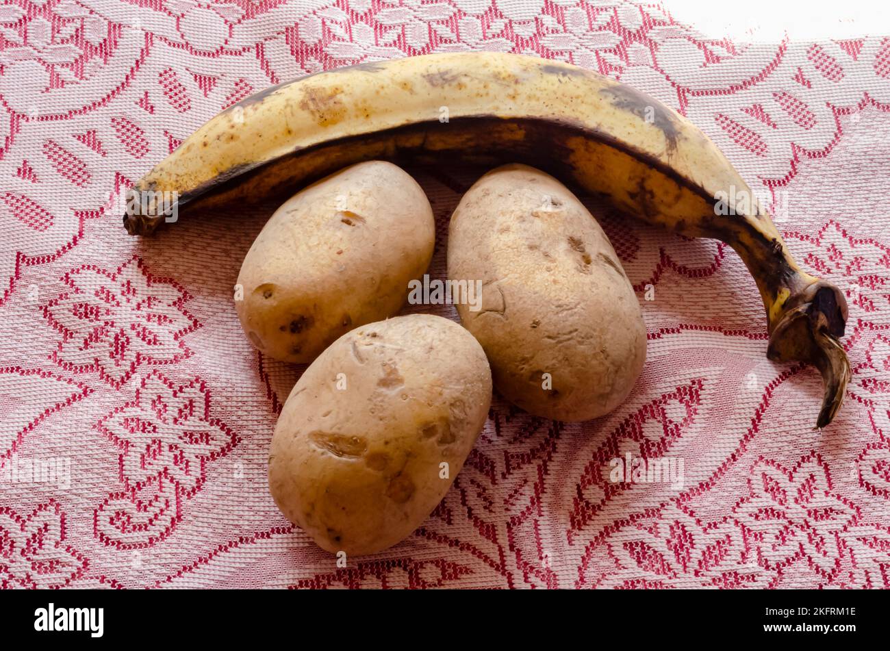 A ripe plantain and three white potatoes are on a red and white lace ...