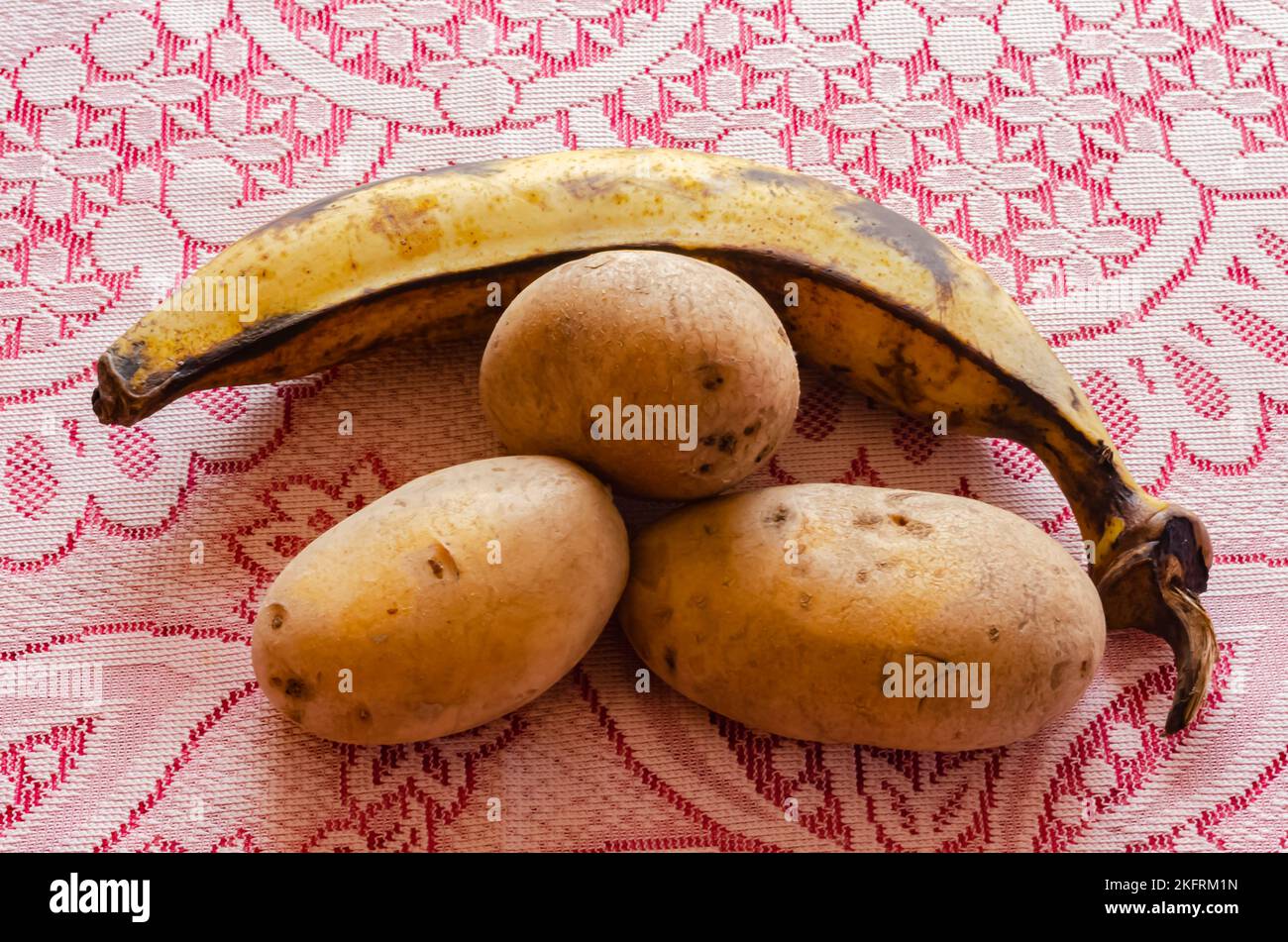 A Plantain and three white potatoes are on a red and white lace ...