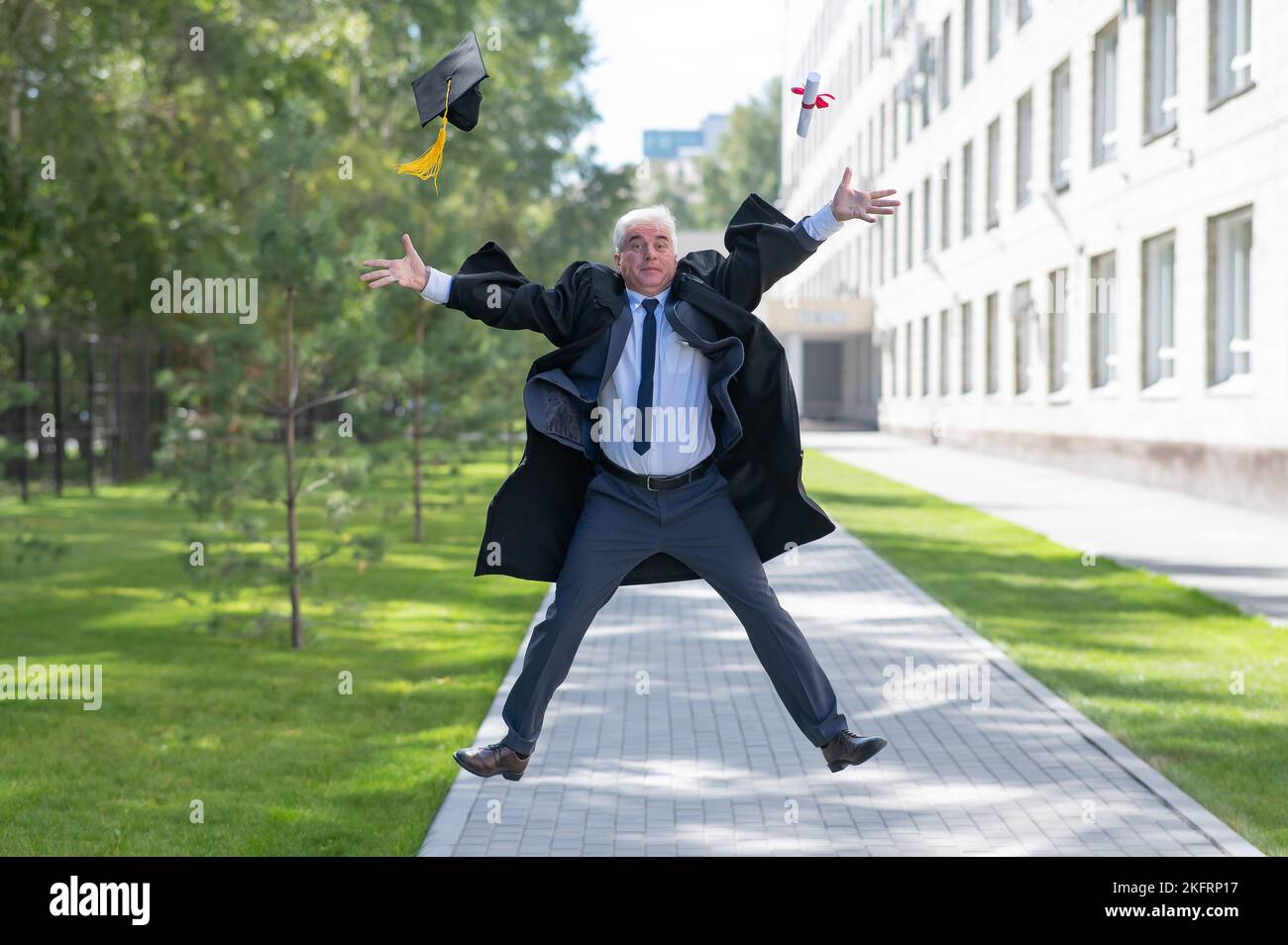Old happy man in graduation gown jumping outdoors and holding diploma ...