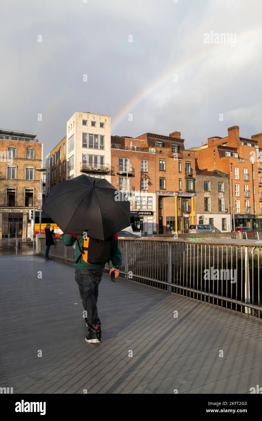 A man crosses a bridge in Dublin underneath an umbrella as a rainbow ...