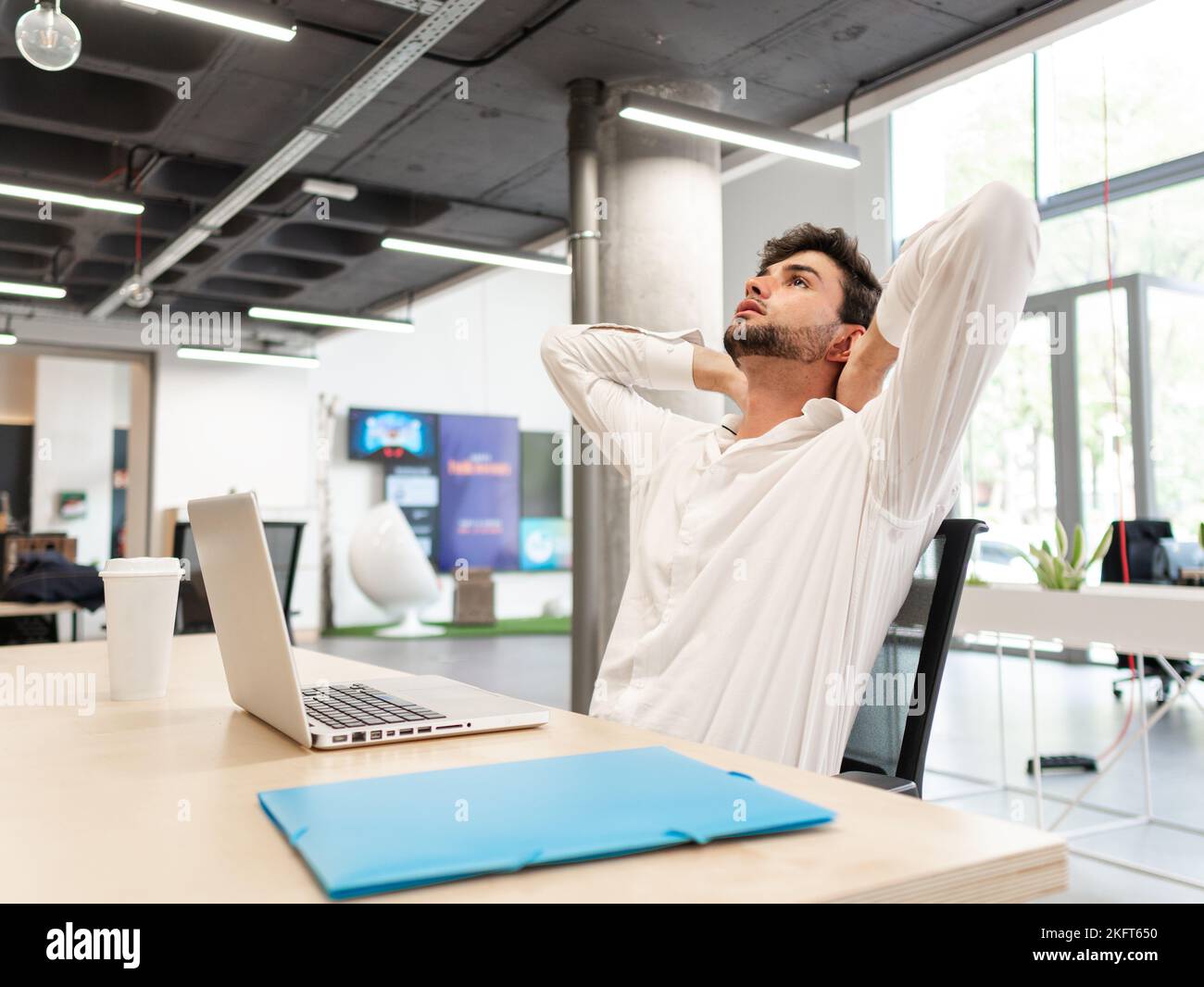 Weary bearded young man in white shirt sitting at table with laptop and ...