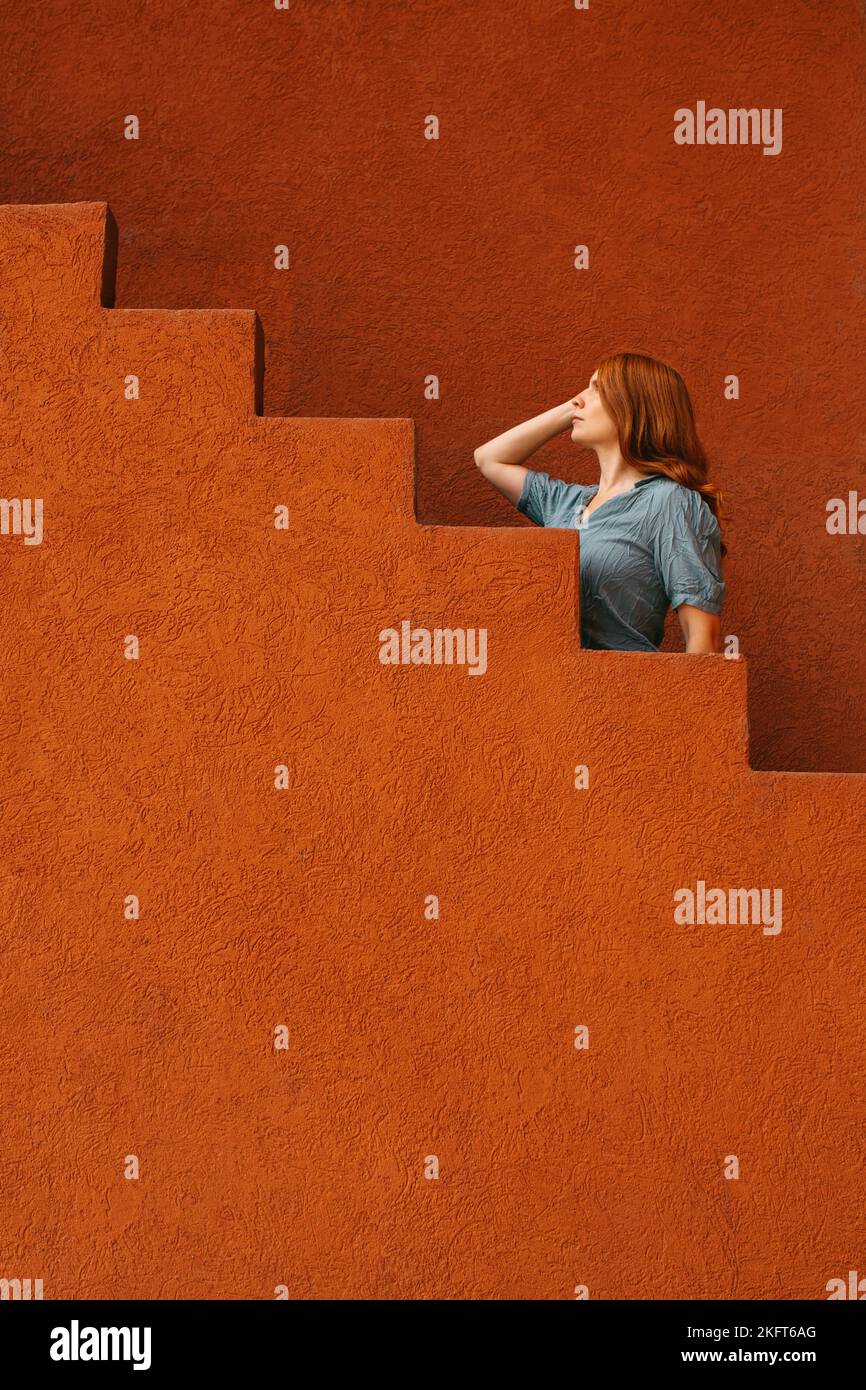 Side view of young redhead female standing in building with orange walls near staircase ...