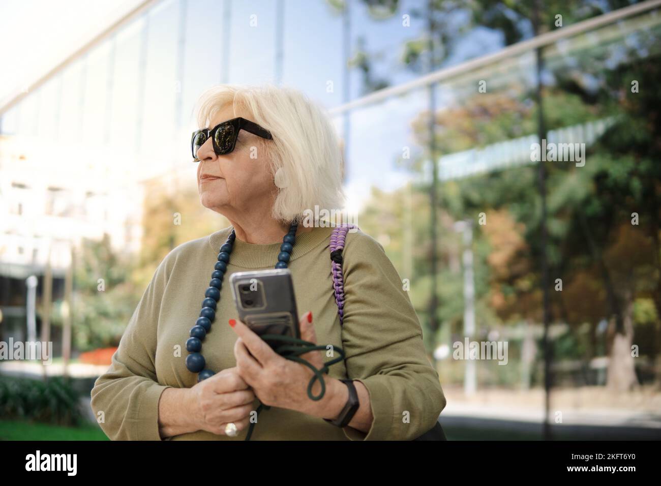 Aged lady in sunglasses and beads browsing on mobile phone standing on ...