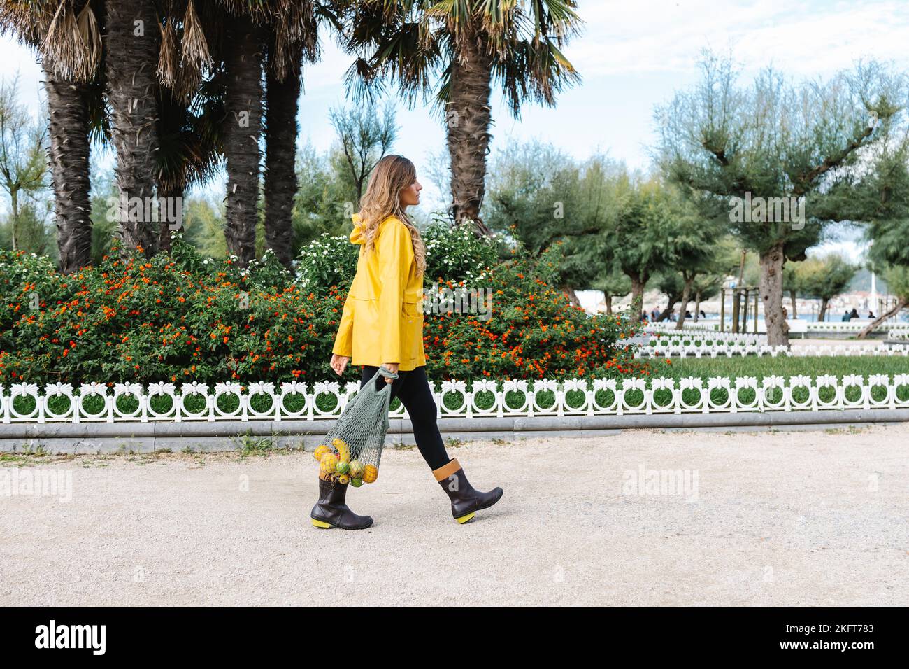 Side view of full body female in bright yellow coat strolling in park ...
