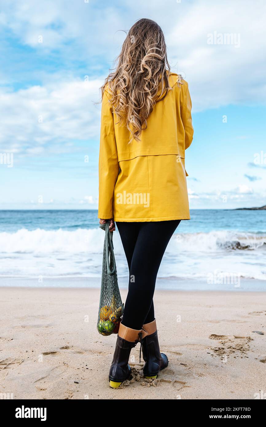Back view of anonymous female in bright yellow coat standing on sandy ...