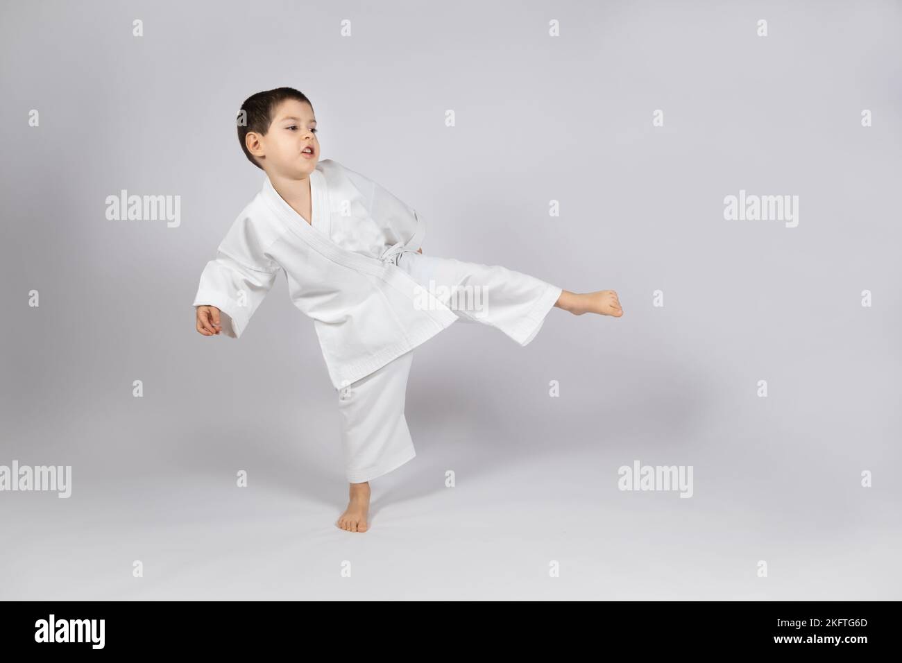 A little boy in a kimono practices karate on a white background ...