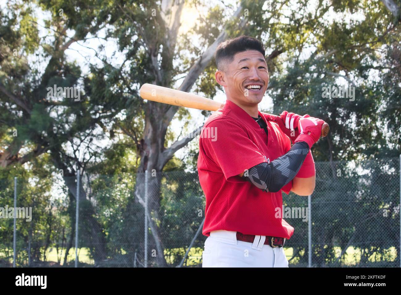 A young Asian man holding a baseball bat standing in a ballpark smiling ...