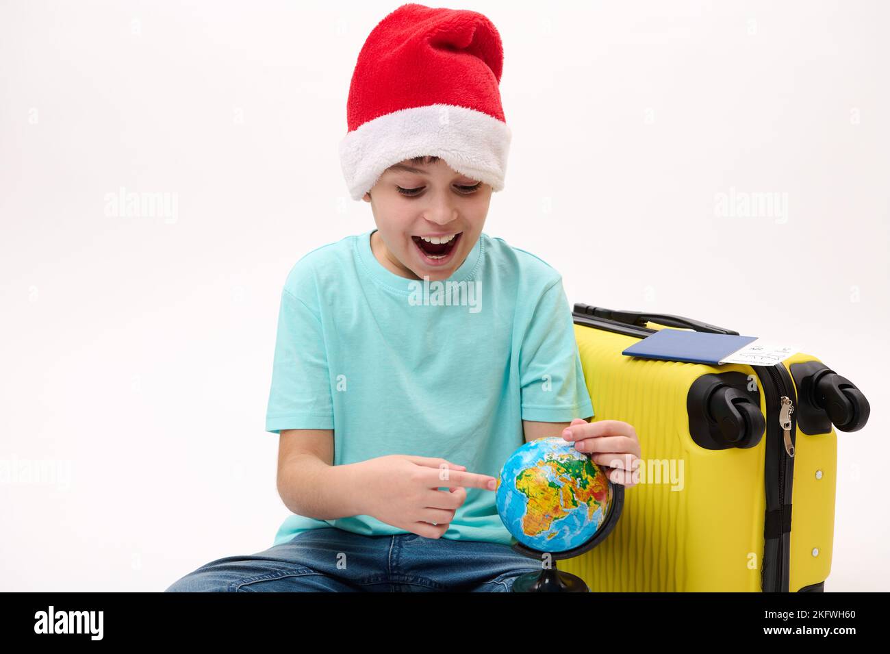 Mischievous teenage boy in Santa hat, sitting next to yellow suitcase ...
