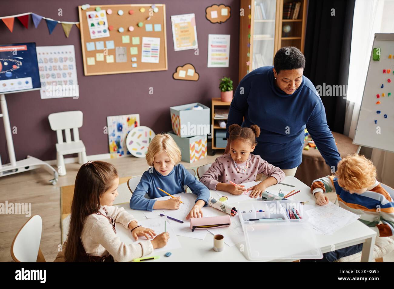 Teacher of drawing bending over boy sitting by table among other little ...