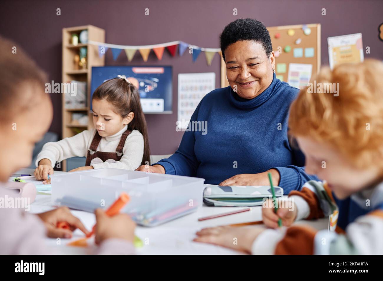 Smiling mature teacher of nursery school looking at one of learners ...