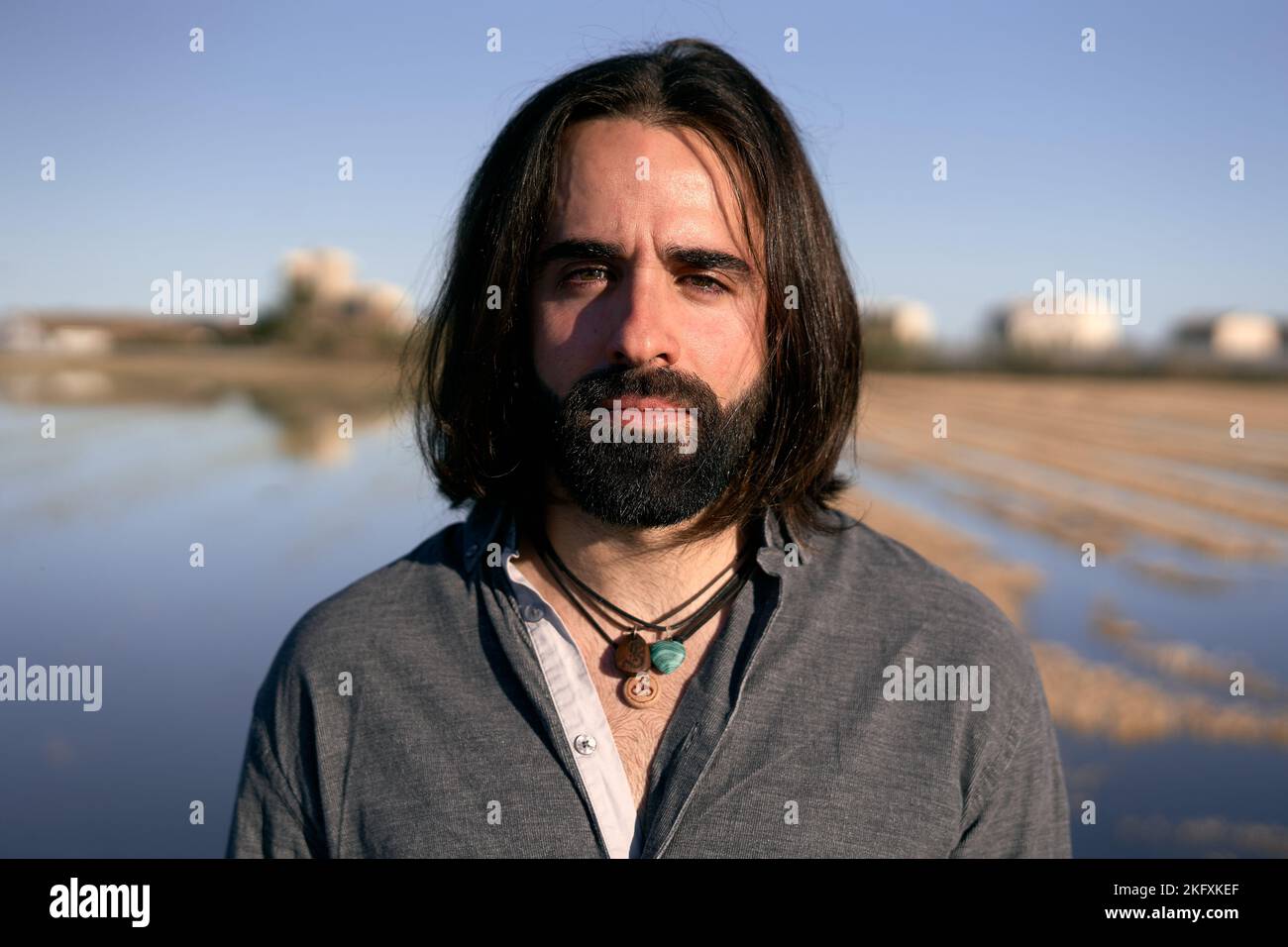 Caucasian young man with beard and long hair standing looking at camera ...