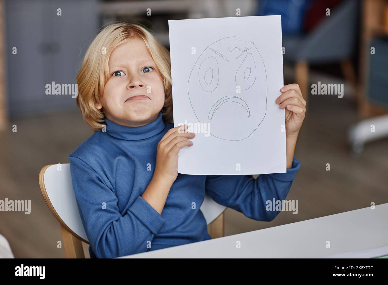 Cute blond boy imitating expression of gloomy face drawn on paper that ...