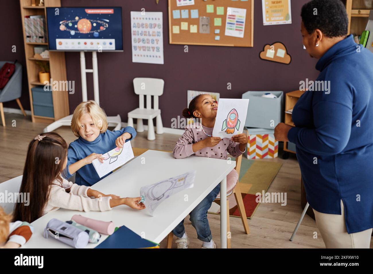 Cute little learner of primary school boasting with her drawing of rocket while showing paper to ...