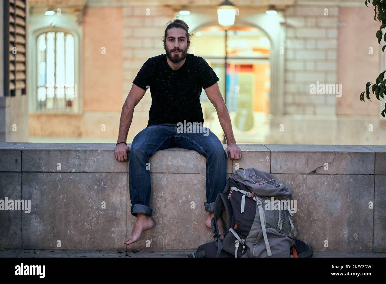 bearded caucasian young man sitting on a wall next to a large backpack ...