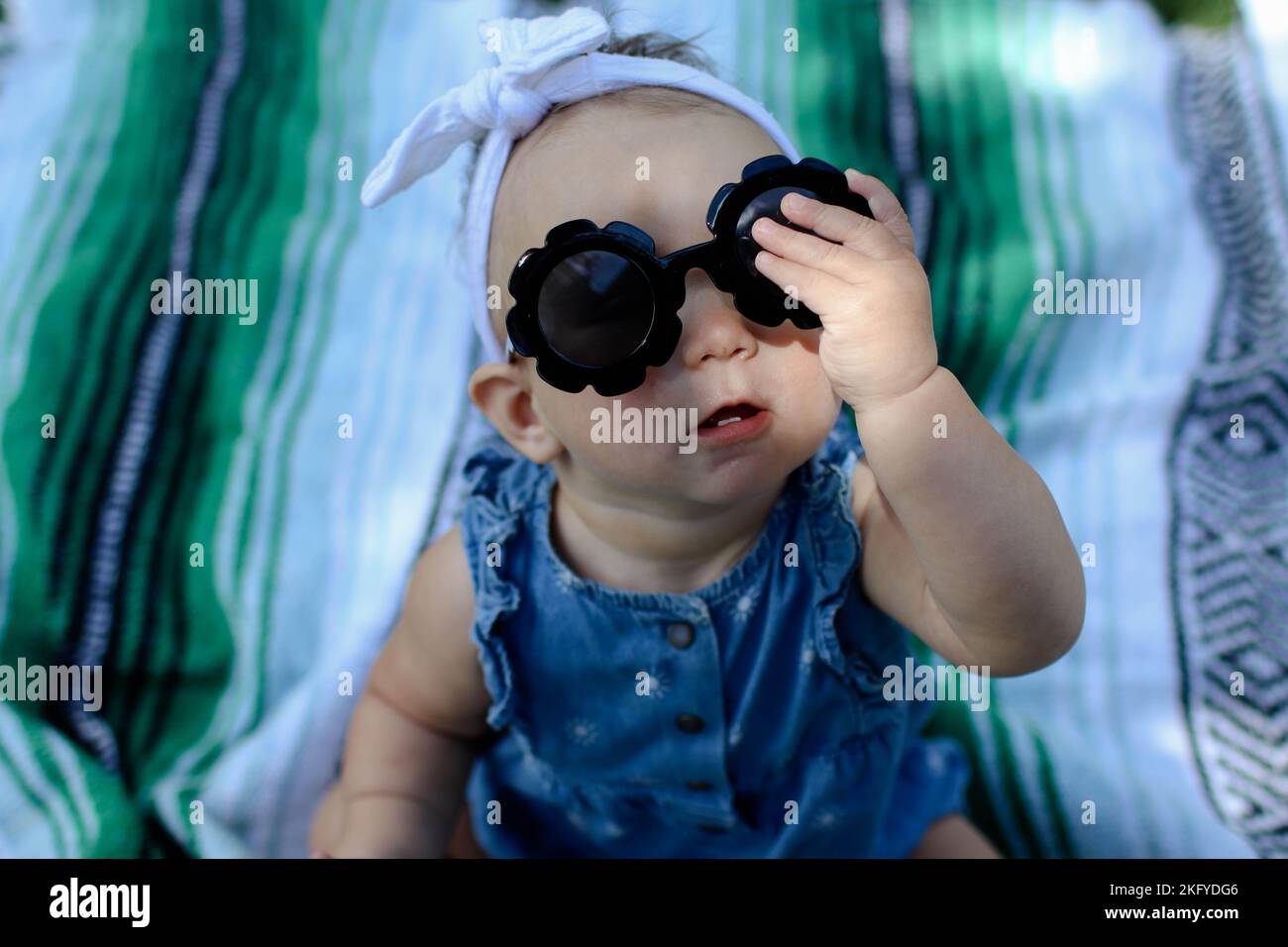 A cute 9-month-old baby sitting on a green picnic blanket with ...