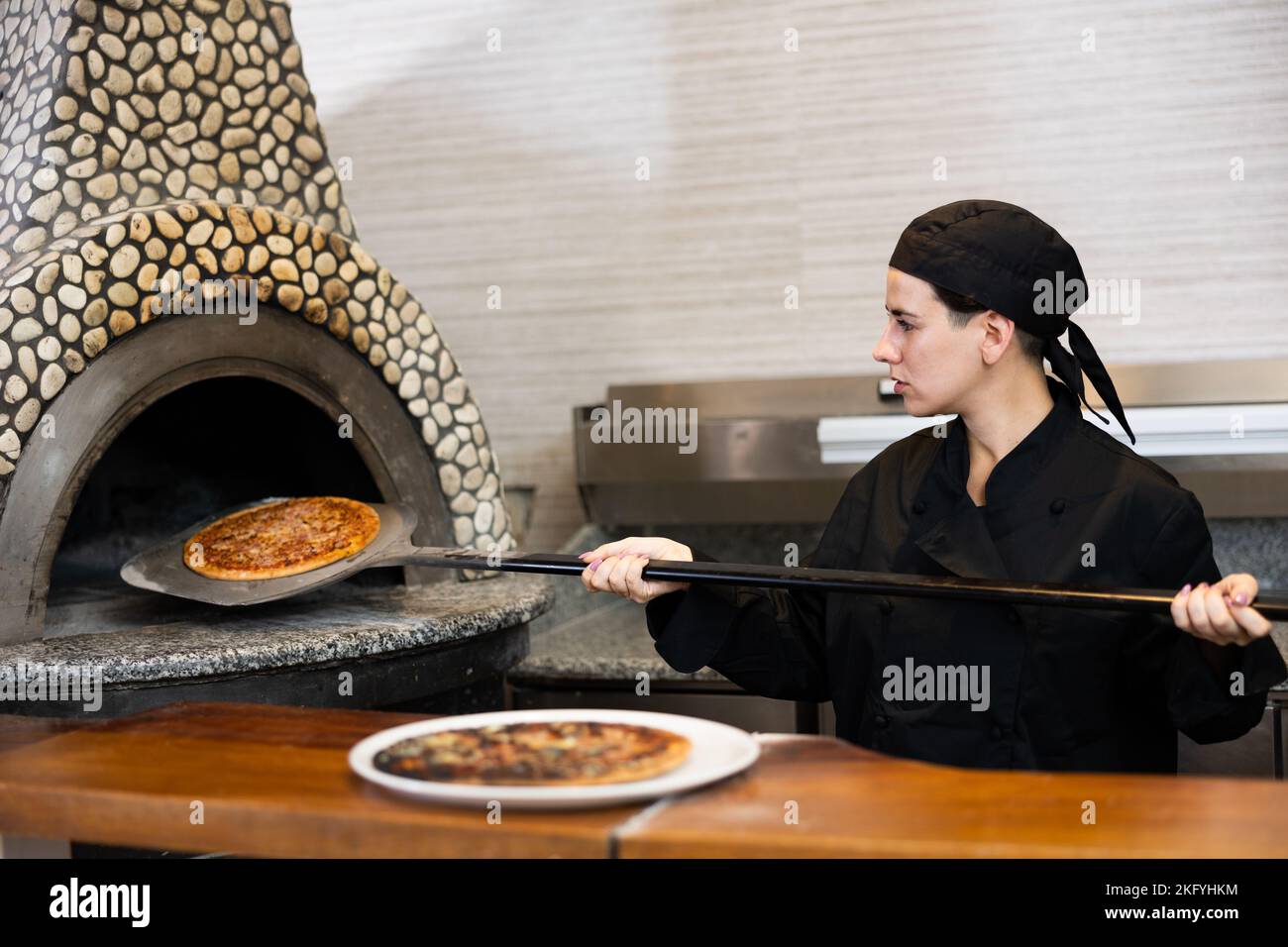 Female chef in black uniform prepares pizza and pulls it out of oven in ...