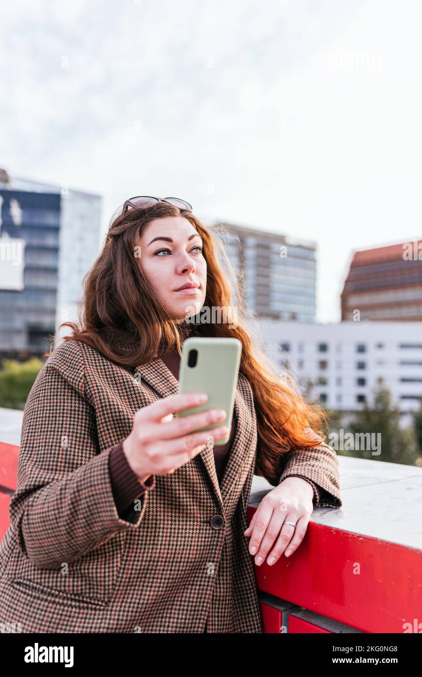 Pensive female in outerwear using cellphone and looking away while ...