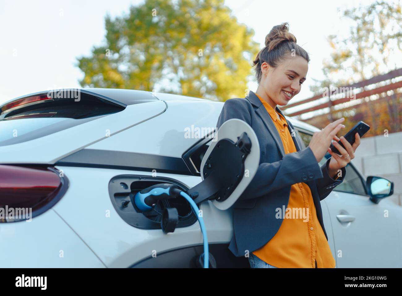 Young woman with smartphone waiting while her electric car charging in ...