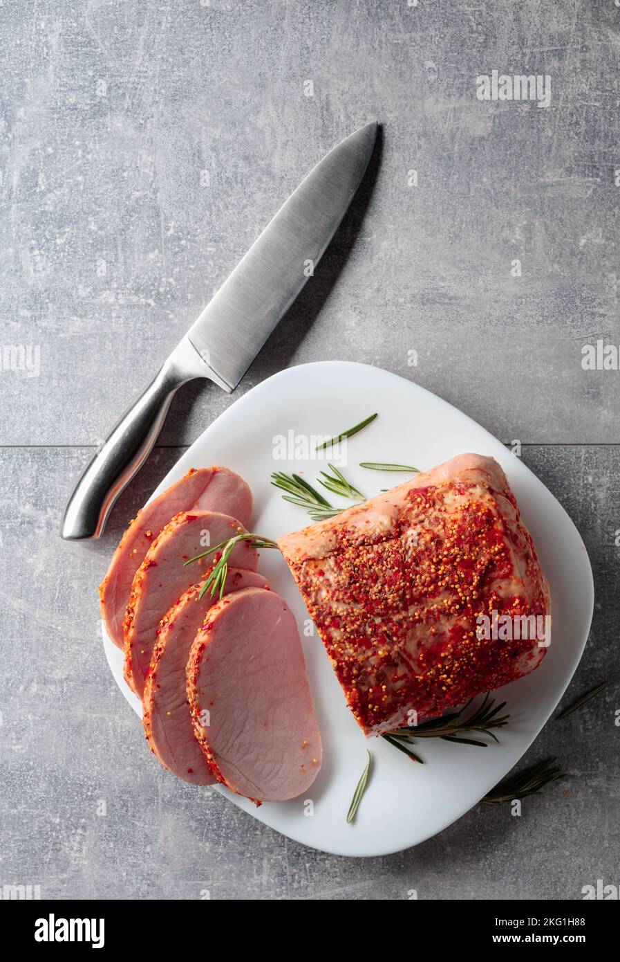 Spicy smoked ham with rosemary and kitchen knife on a grey stone table ...