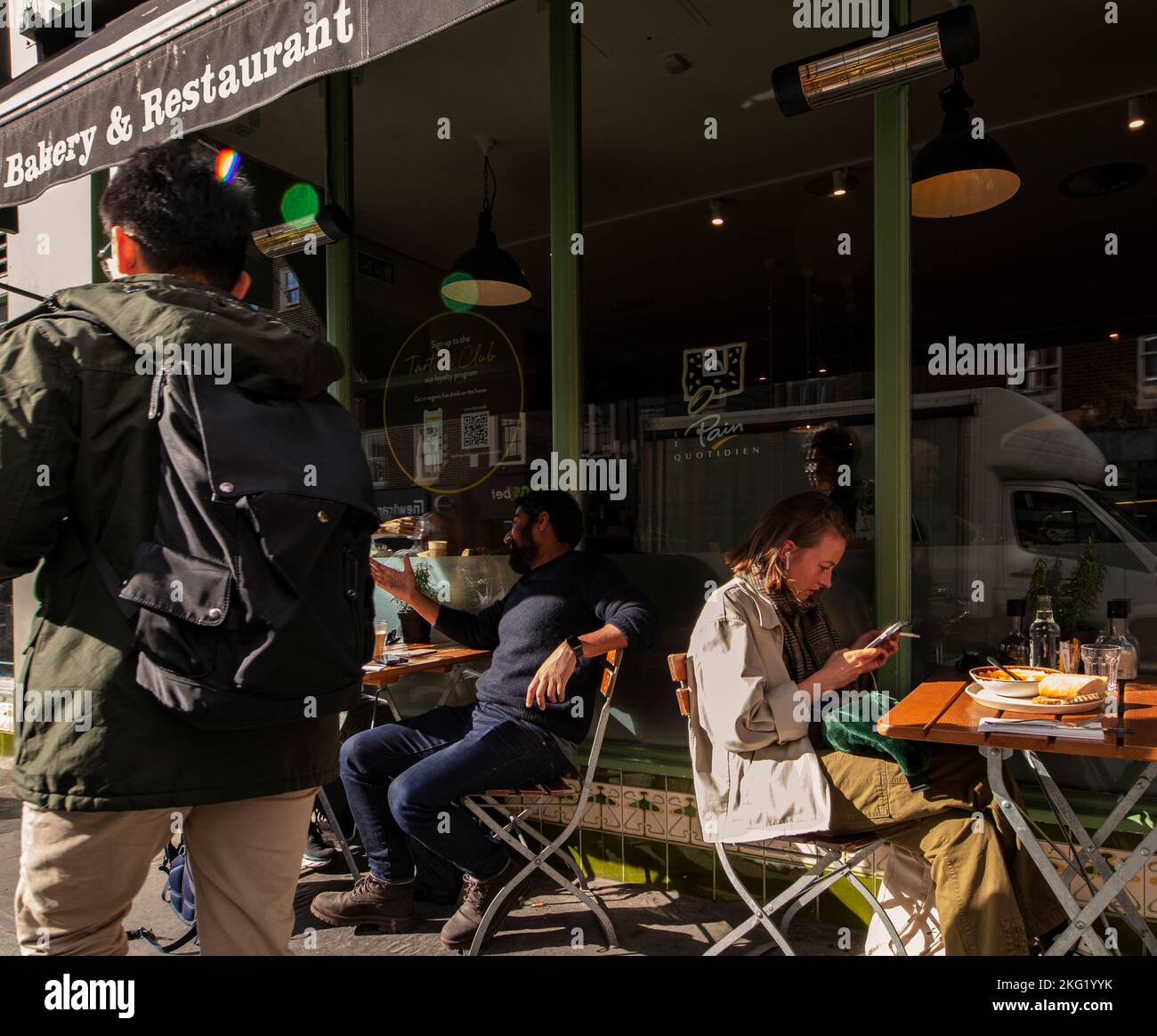 Exterior of restaurant in London, people sitting on tables outside in the street, checking ...