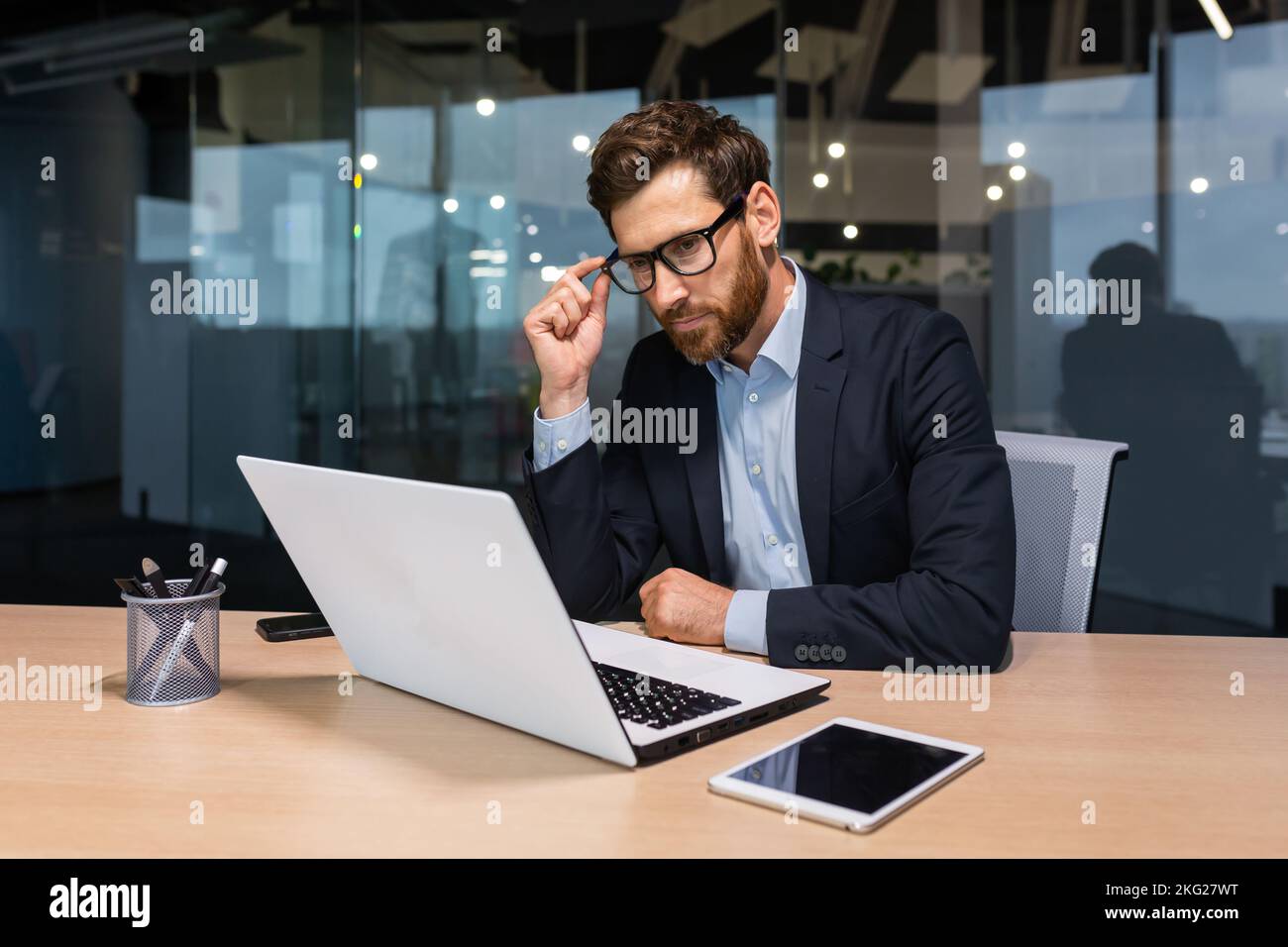 Serious thinking senior businessman working inside office using laptop, man in glasses solving ...