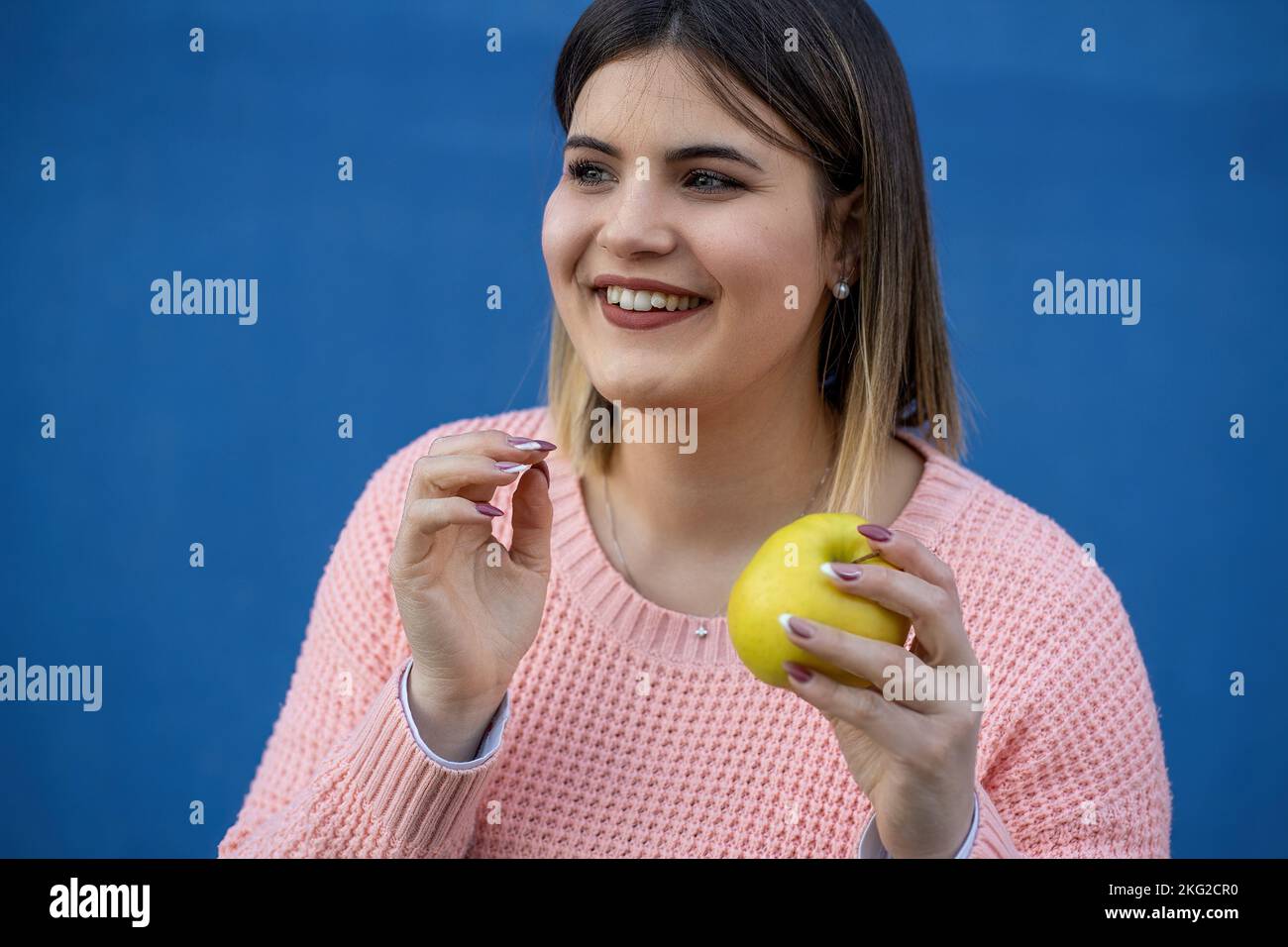 Cheerful chubby woman holding a yellow apple, isolated against a blue ...