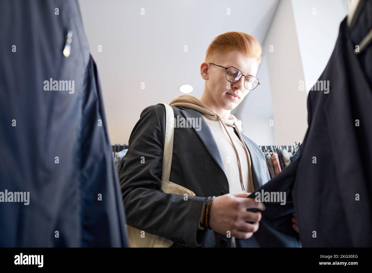 Low angle portrait of red haired young man browsing clothes on racks ...