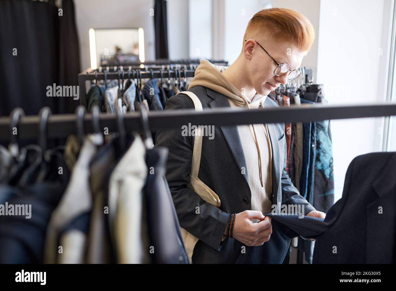 Side view of young red haired man browsing clothes on racks while ...