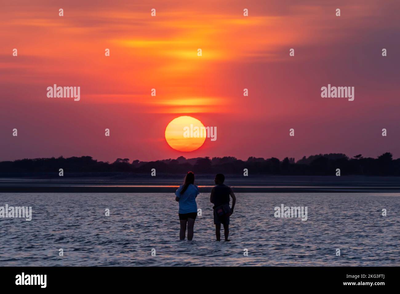 West Wittering sunset with a couple standing in the shallow water watching the sun go down ...