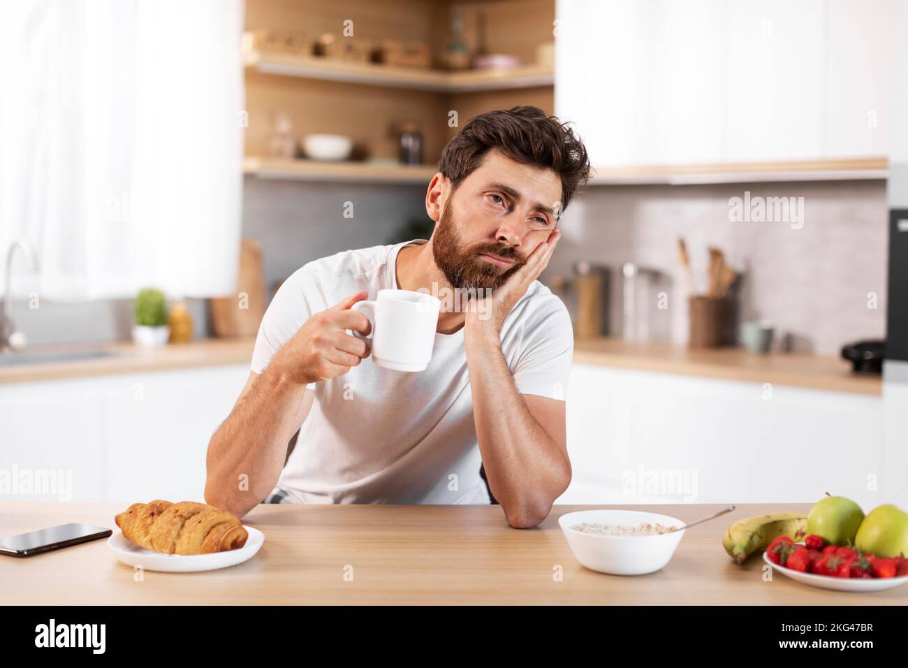Sad tired middle aged european bearded male holds cup, drinks coffee ...