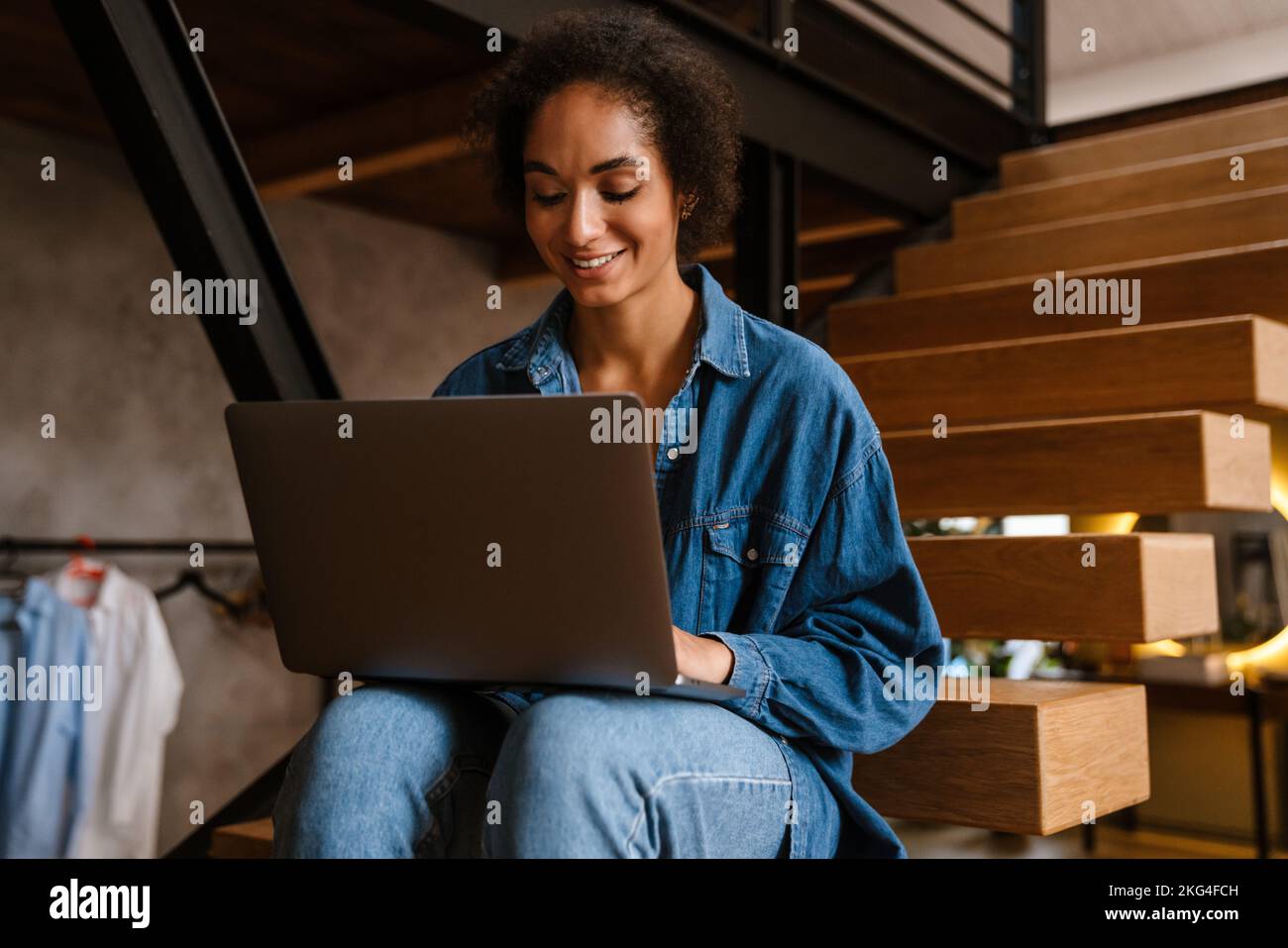 Young black woman wearing denim shirt smiling and using laptop at home ...