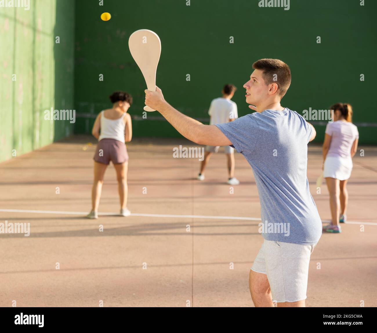 Guy playing pelota at open-air fronton, swinging wooden bat to return ...