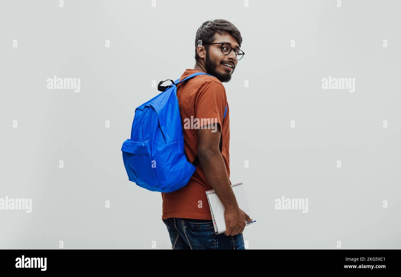 Indian student with blue backpack, glasses and notebook posing on gray ...