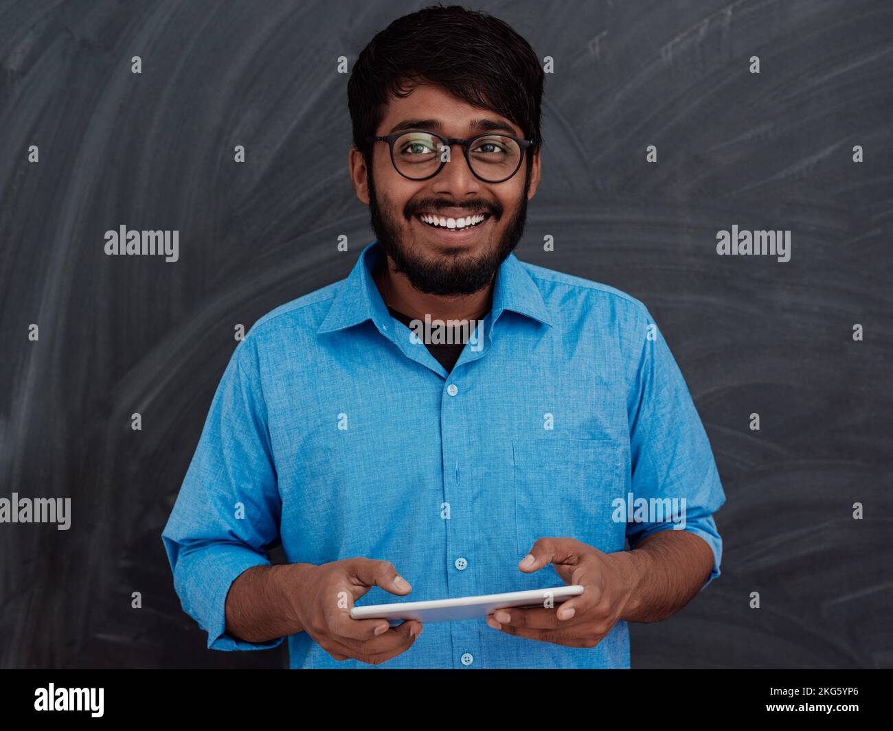 Indian smiling young student in blue shirt and glasses using tablet ...