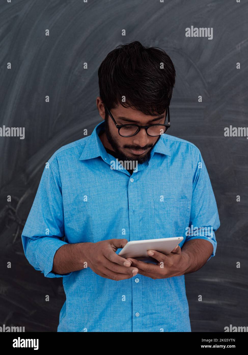 Indian smiling young student in blue shirt and glasses using tablet ...