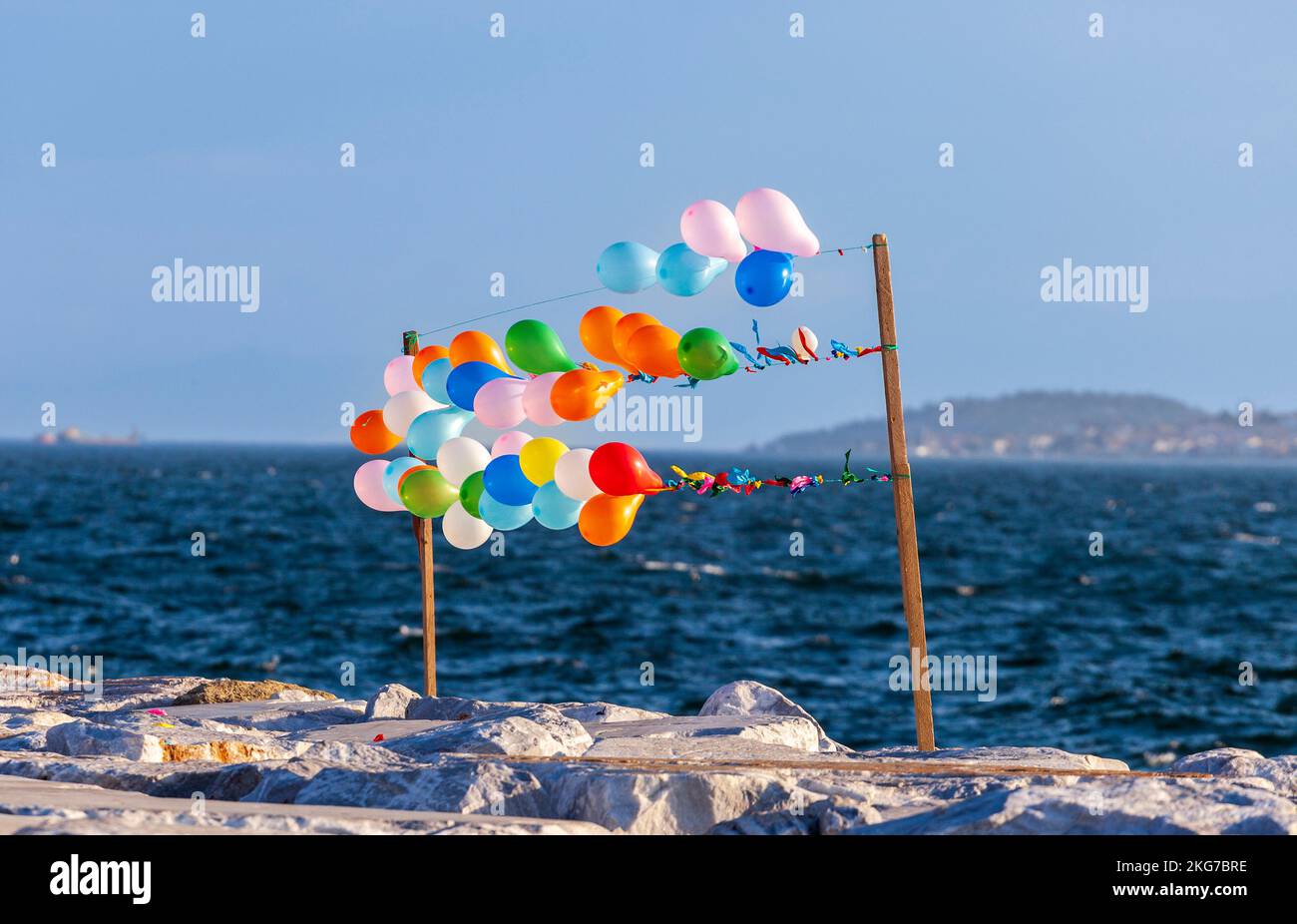 Colorful balloons tied to poles on a rocky shore by the sea, creating a ...