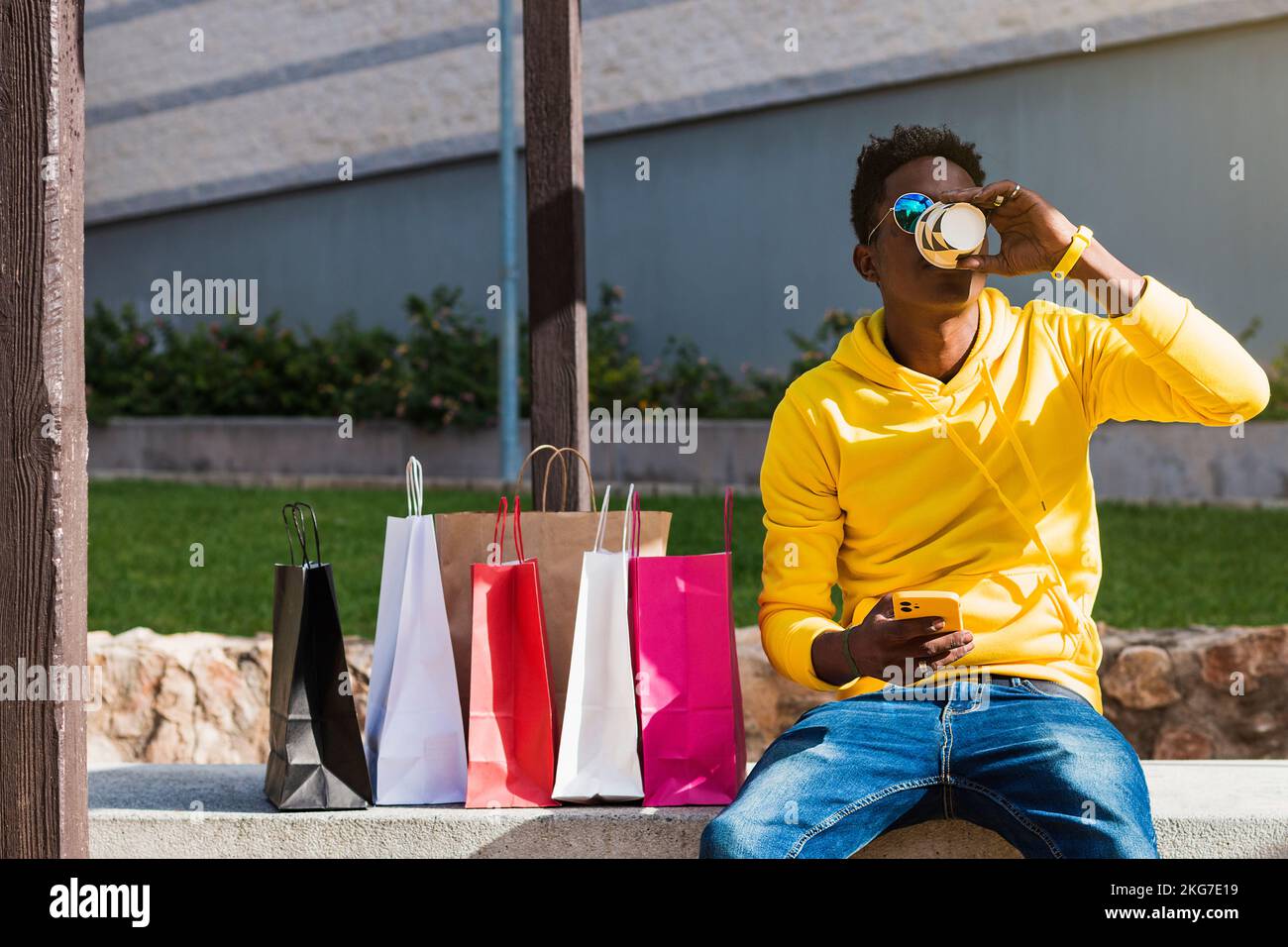 Young African man sitting on a park bench drinking coffee from a ...