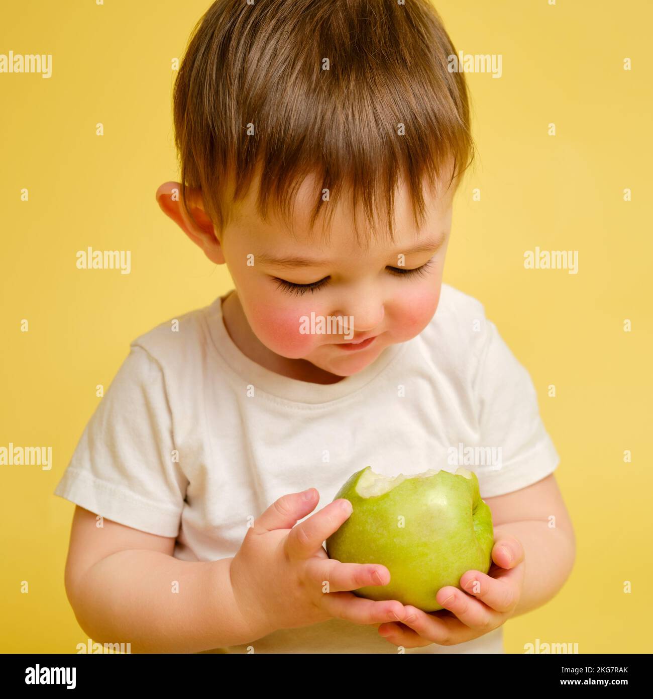 Happy toddler baby eating a green apple on studio yellow background ...