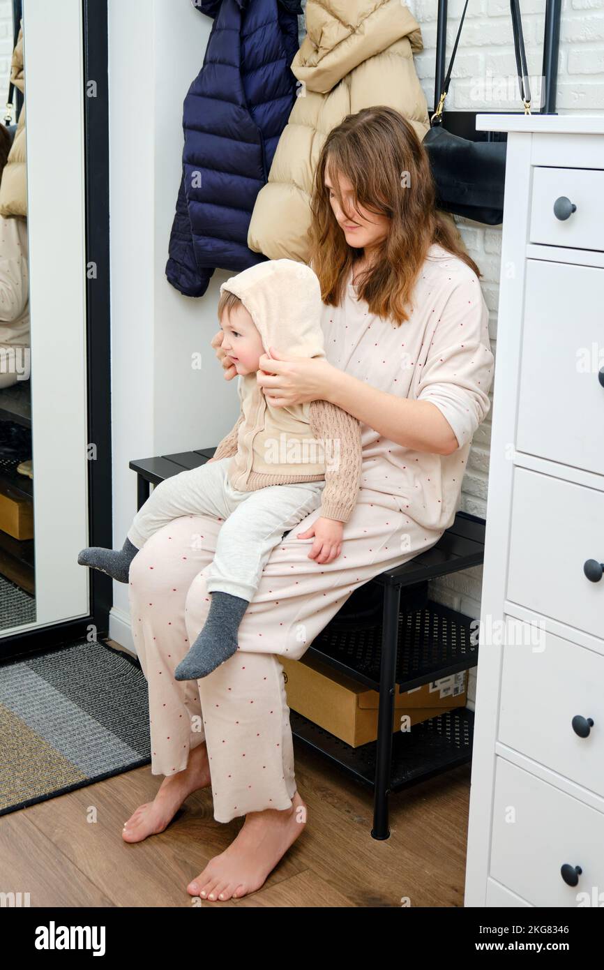 A mother puts a hoodie jacket on a toddler baby sitting in the home ...