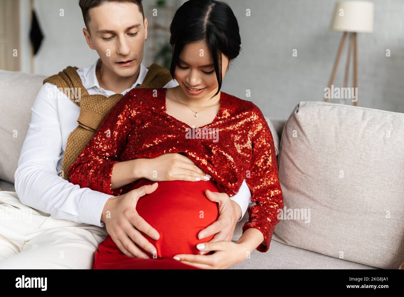 young man embracing belly of pregnant asian wife wearing red and shiny ...