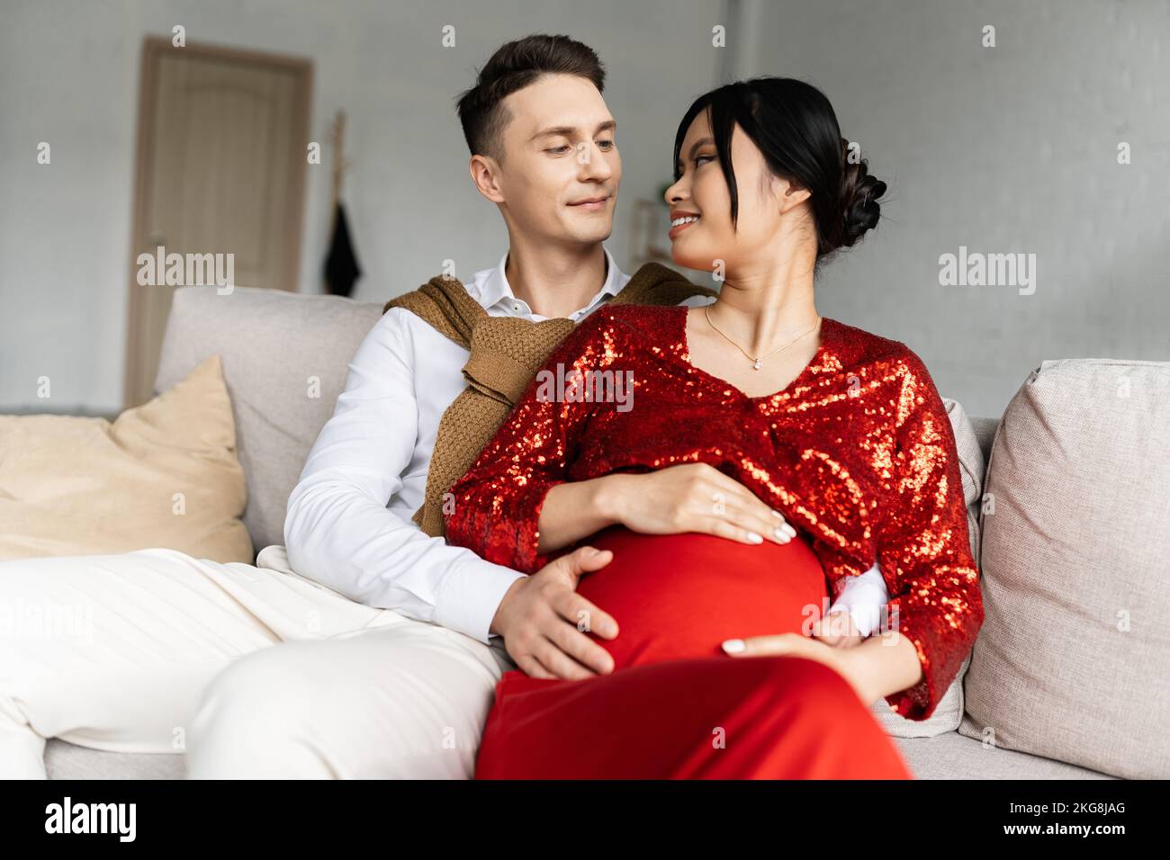 Pregnant Asian Woman In Red And Shiny Clothes Smiling At Young Husband Hugging Her Tummy On