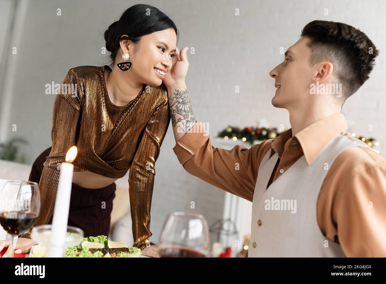 tattooed man stroking hair of asian wife near festive dinner on dining ...