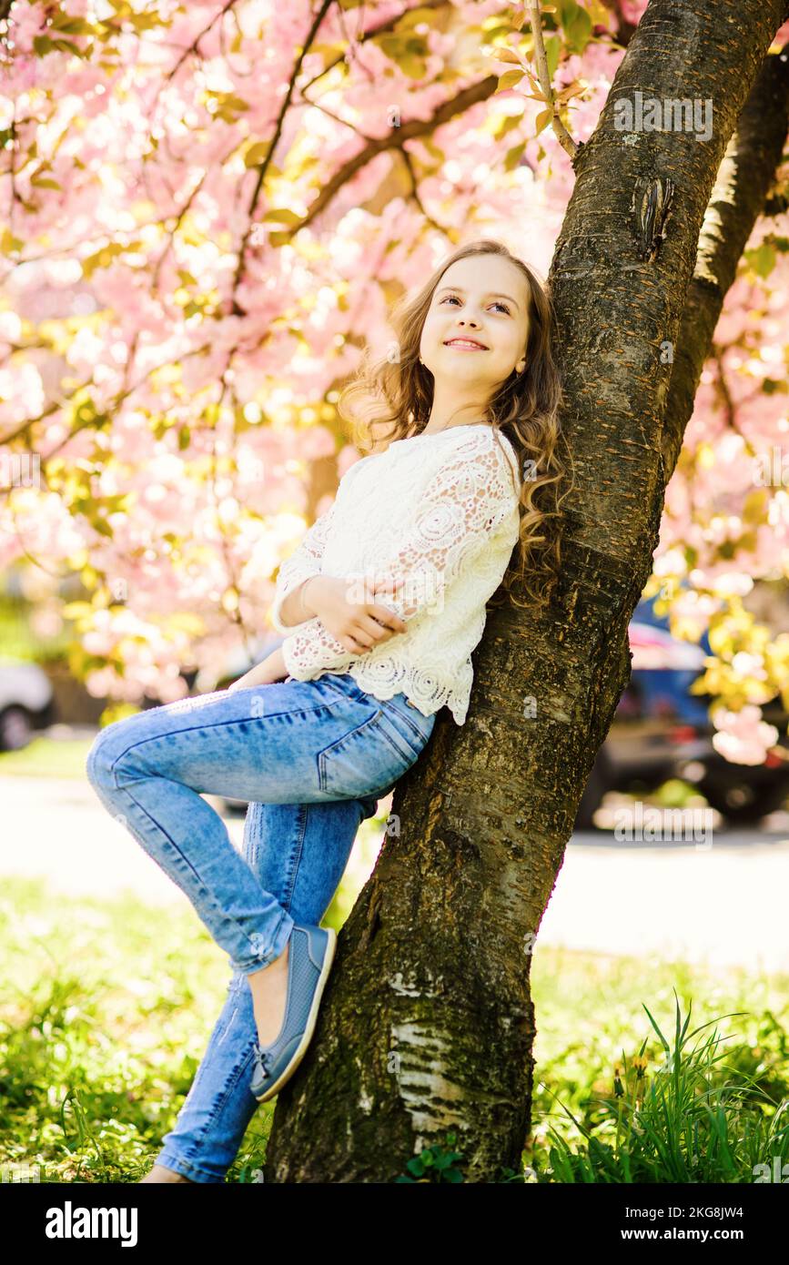 Girl on smiling face standing near sakura, lean on tree trunk. Girl ...