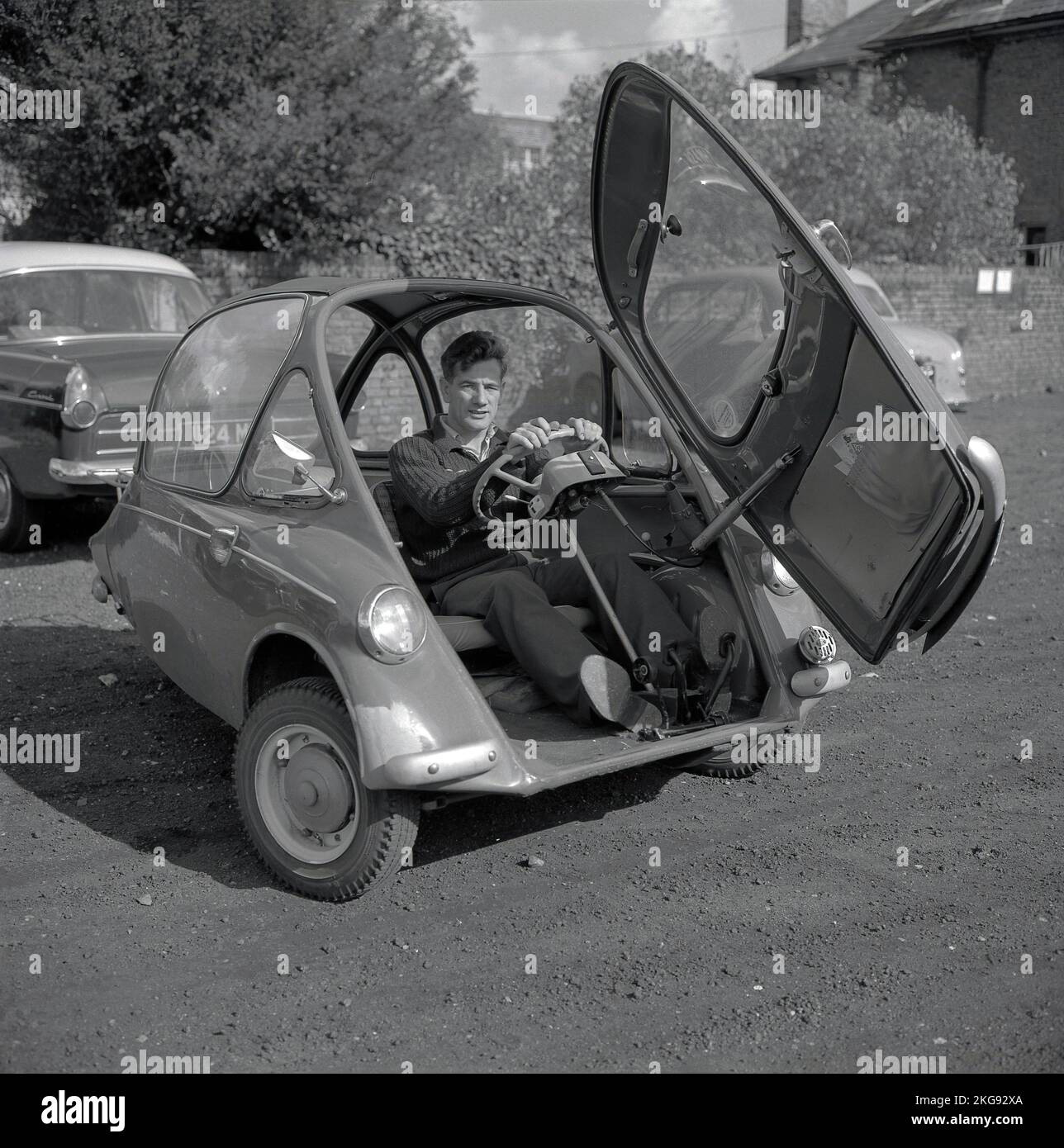 1960s, historical, outside in a gravel car park, a man sitting inside a small car of the era, a ...