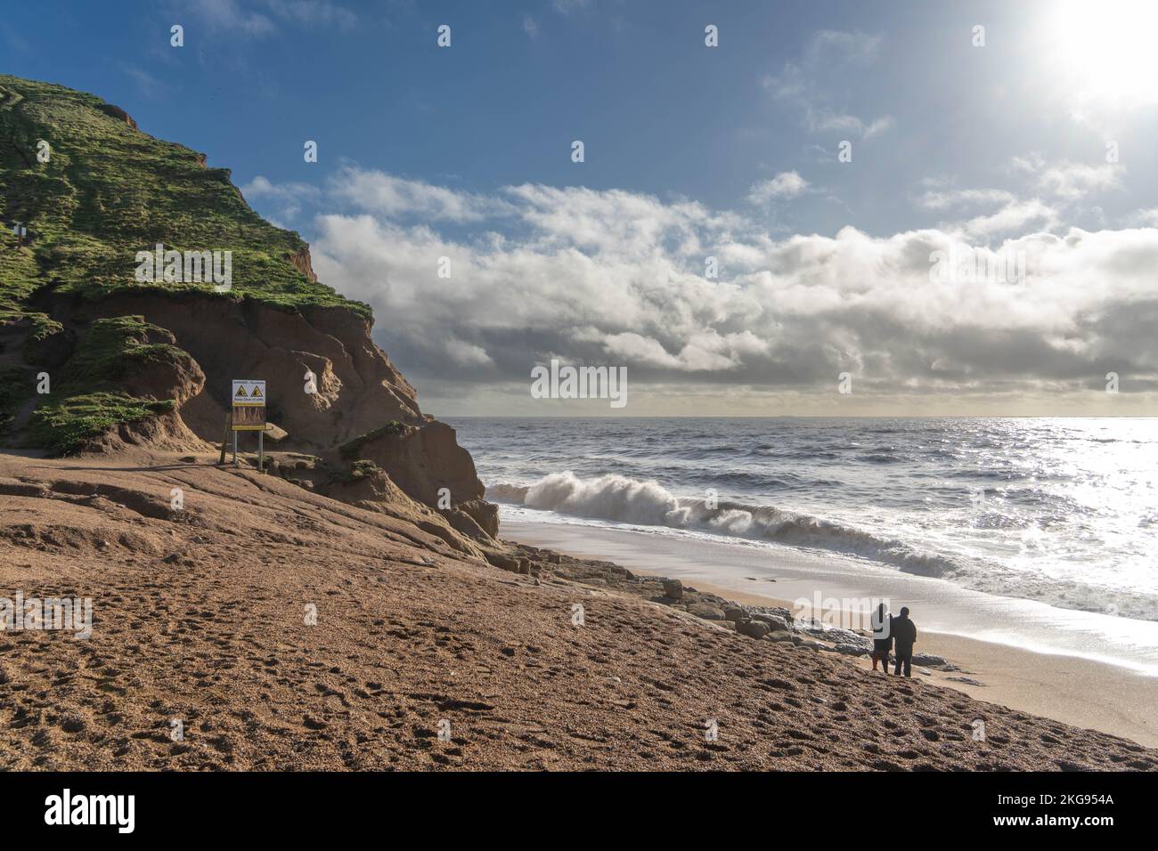 A couple stand on the beach by dramatic sunlit cliffs at West Bay ...