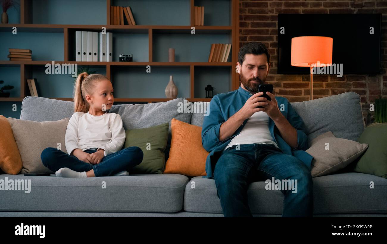 Bored child girl resting at home with father young dad using mobile phone little kid need ...